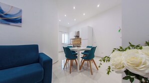 Uma sala moderna com um sofá azul, um espaço de refeições com cadeiras verde-água e uma cozinha ao fundo.