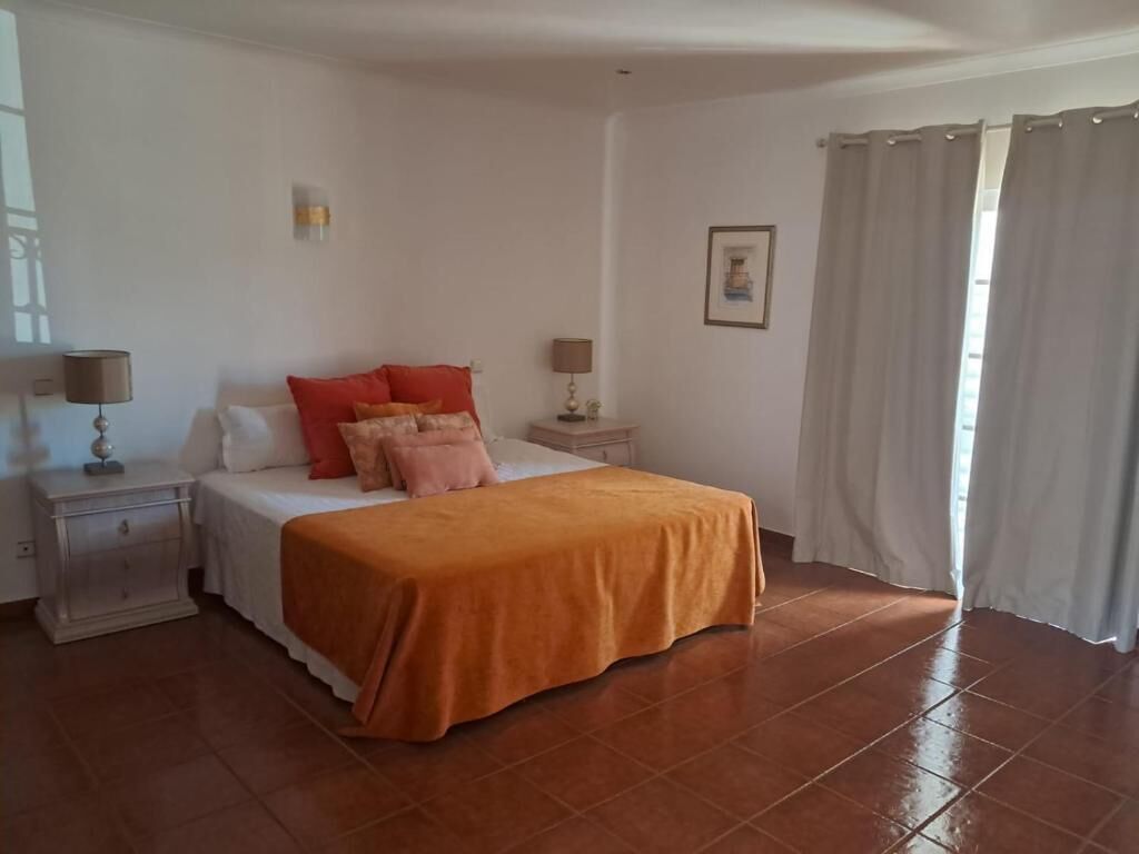 Quarto com cama, mesas de cabeceira, abajur, quadro na parede e janela com cortinas.