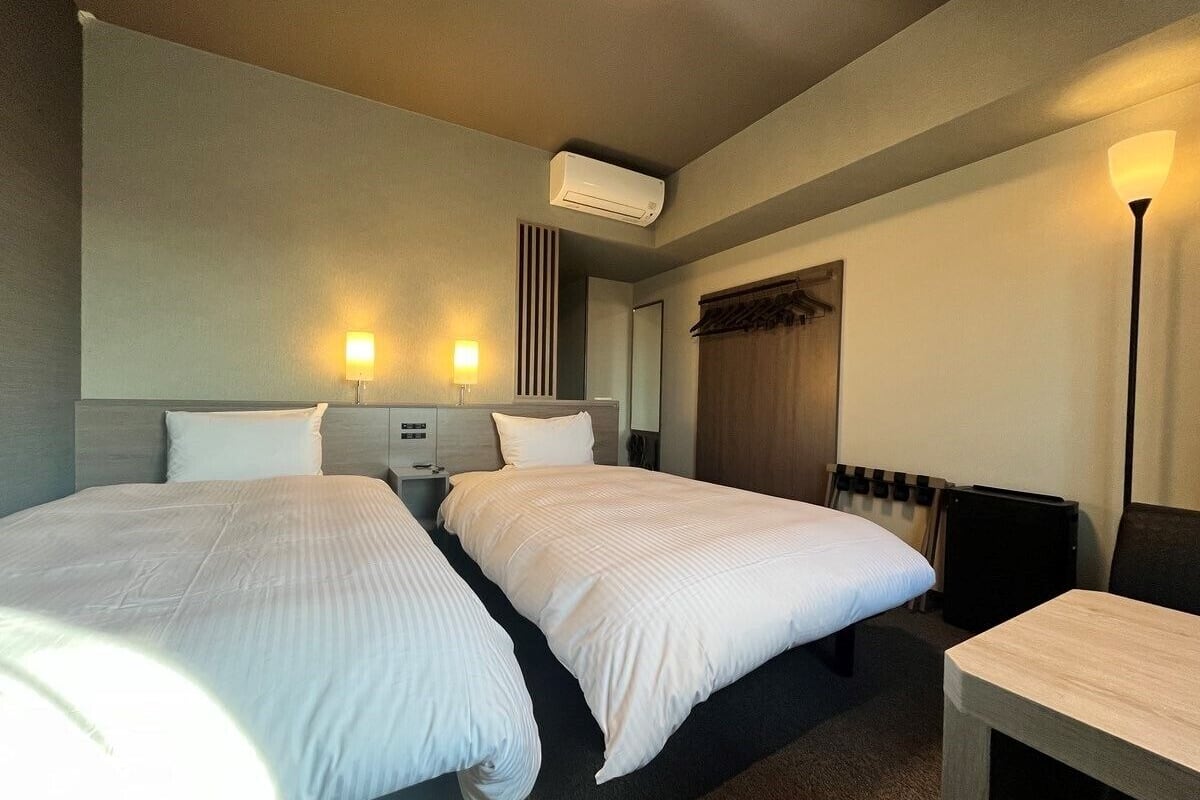 Quarto de hotel com duas camas, uma escrivaninha, uma lâmpada, ar condicionado e radiador.