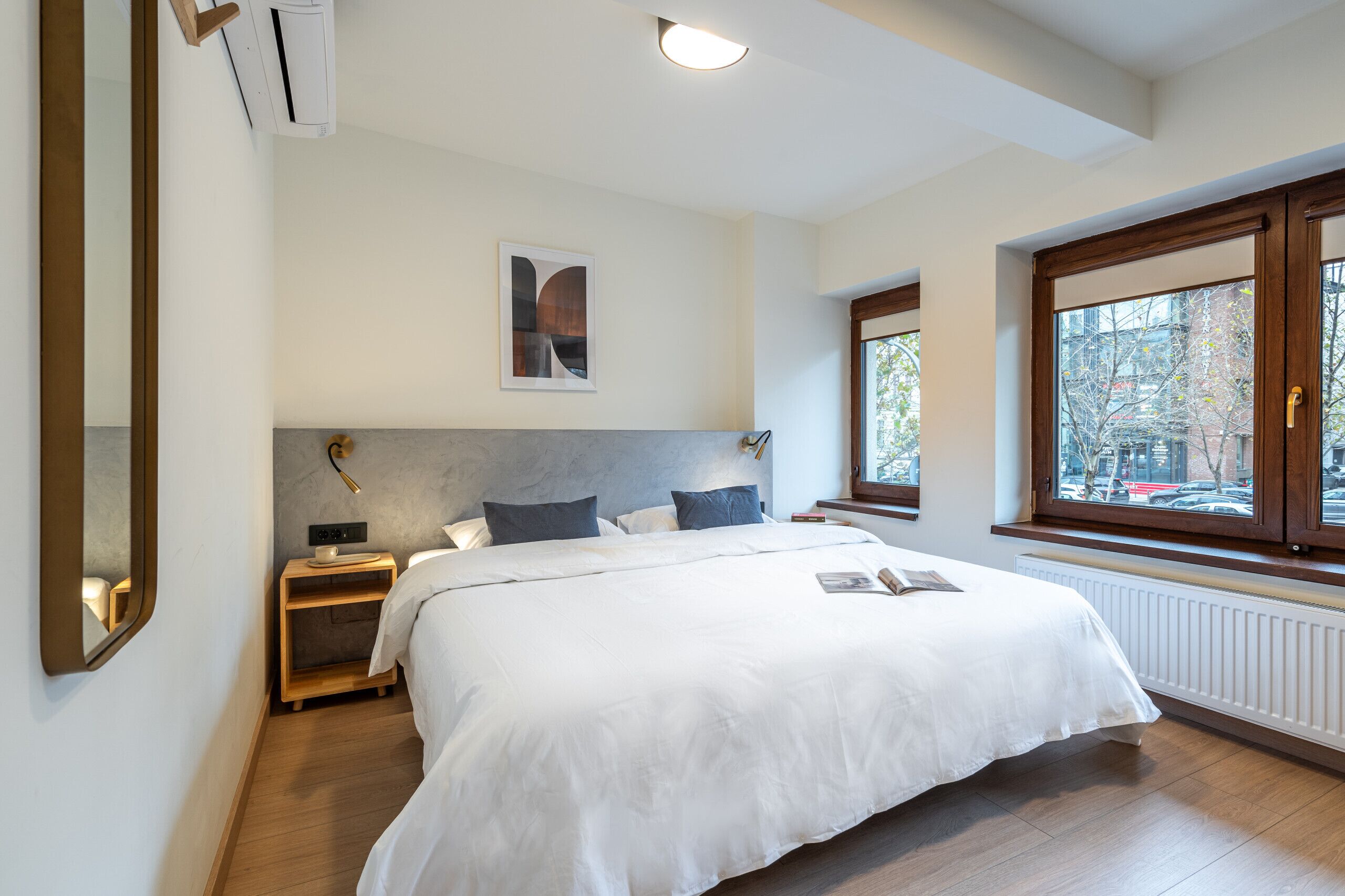 Quarto com cama grande, mesas de cabeceira, criado-mudo, janela com vista para o exterior e uma obra de arte na parede.