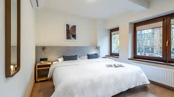 Quarto com cama grande, mesas de cabeceira, criado-mudo, janela com vista para o exterior e uma obra de arte na parede.