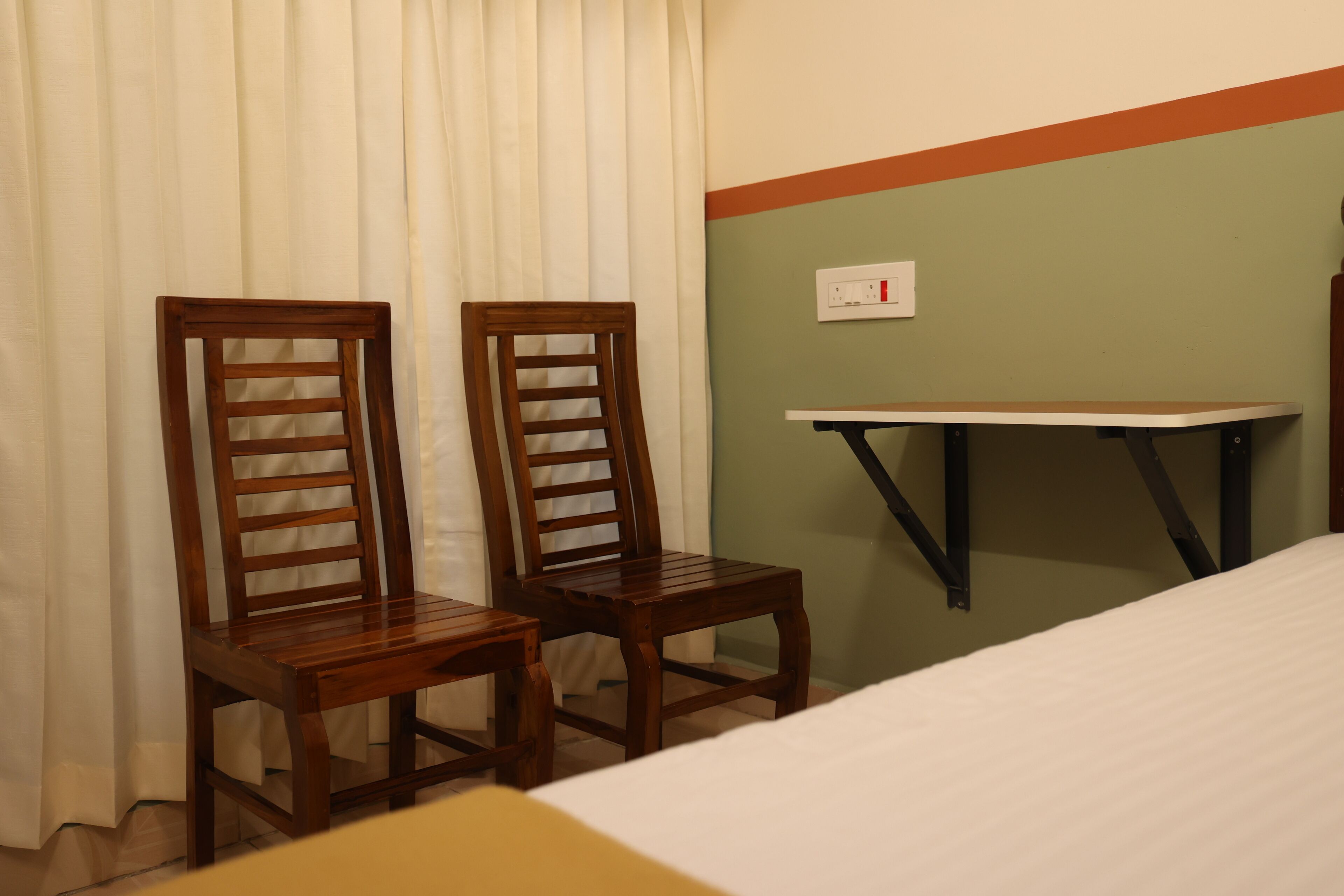 Quarto com duas cadeiras de madeira, uma mesa pequena, uma cama e uma tomada.