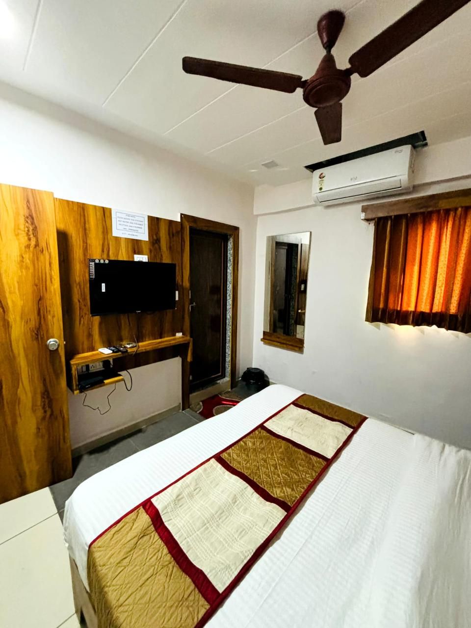 Quarto de hotel com cama, ventilador de teto, ar condicionado, televisão presa a um painel de madeira, janela com cortinas e uma pequena mesa com uma cadeira.