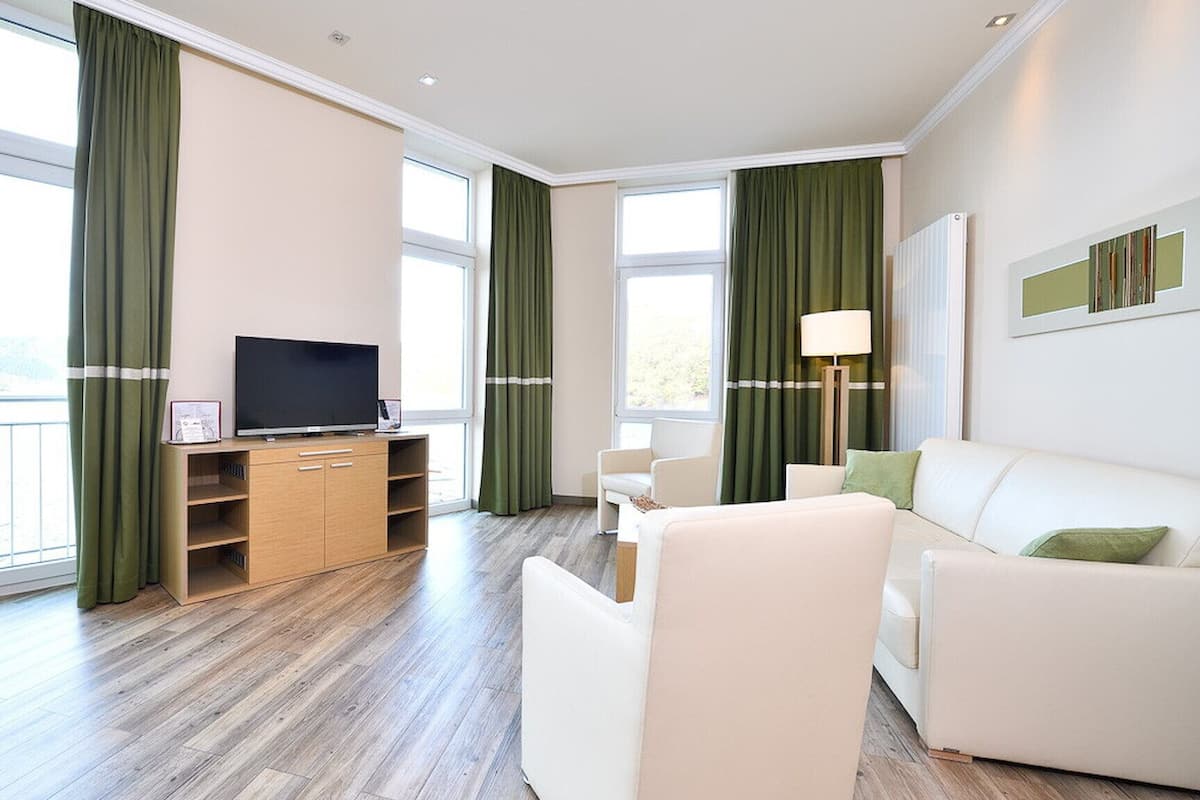 Quarto de hotel moderno com uma grande janela, uma televisão de tela plana sobre um móvel de madeira, um sofá branco e um abajur.