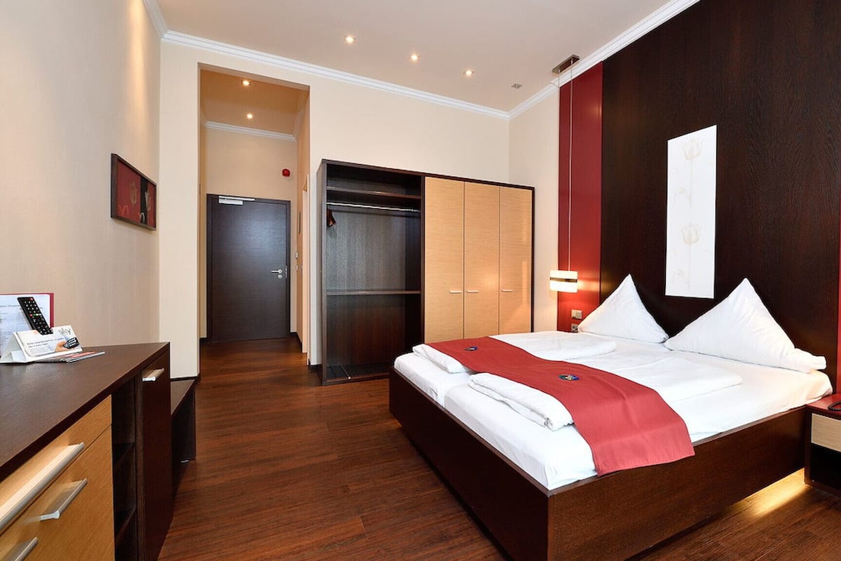 Quarto de hotel com cama, mesa de cabeceira, luminária, guarda-roupa e escrivaninha com telefone.