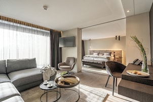 Quarto de hotel moderno com sofá, poltrona, mesa de centro e área de jantar, incluindo um vaso e um prato.