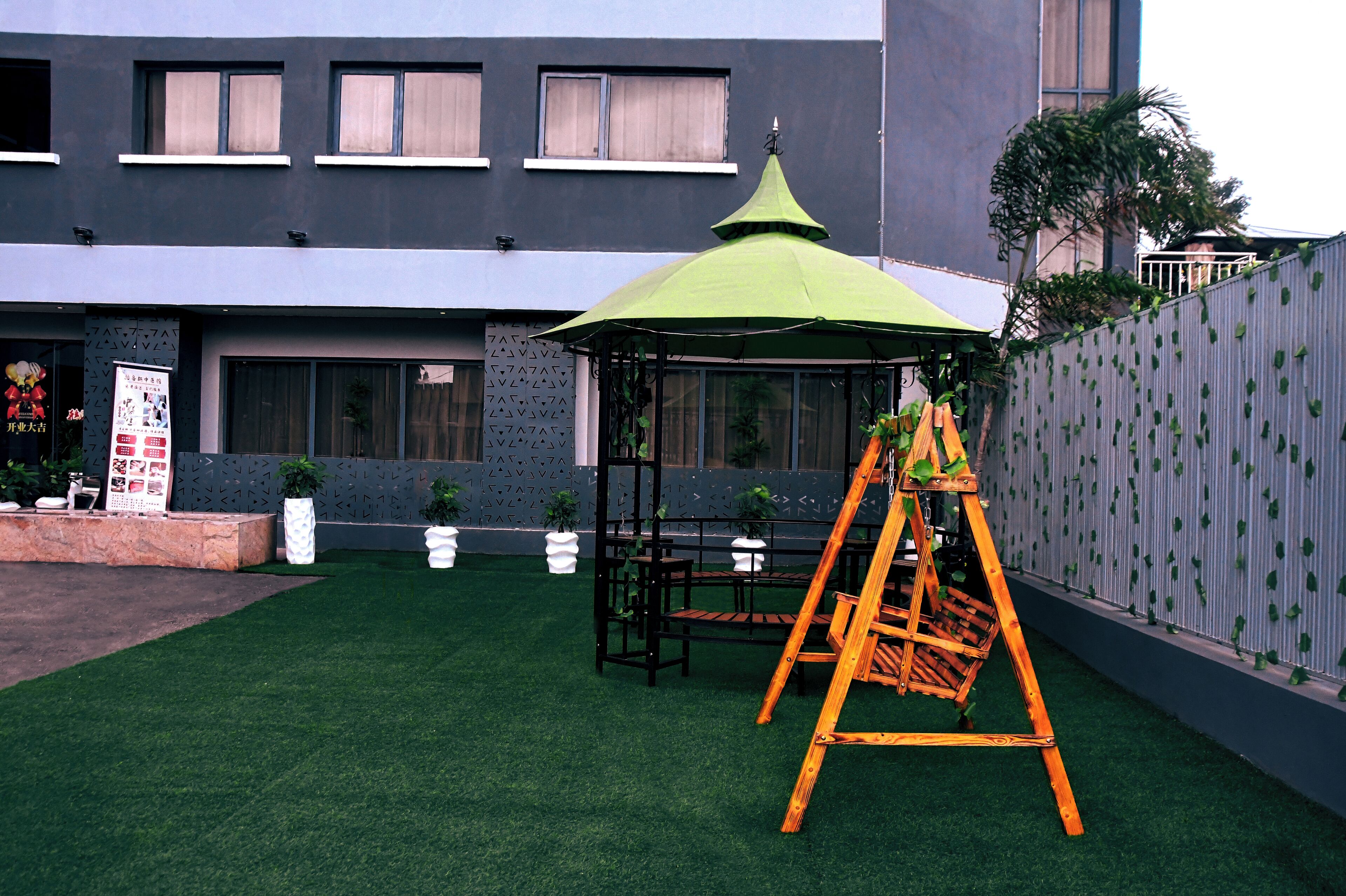 Um pátio com grama sintética, balanço de madeira, um gazebo com telhado verde e um edifício com janelas amplas e fachada cinzenta.