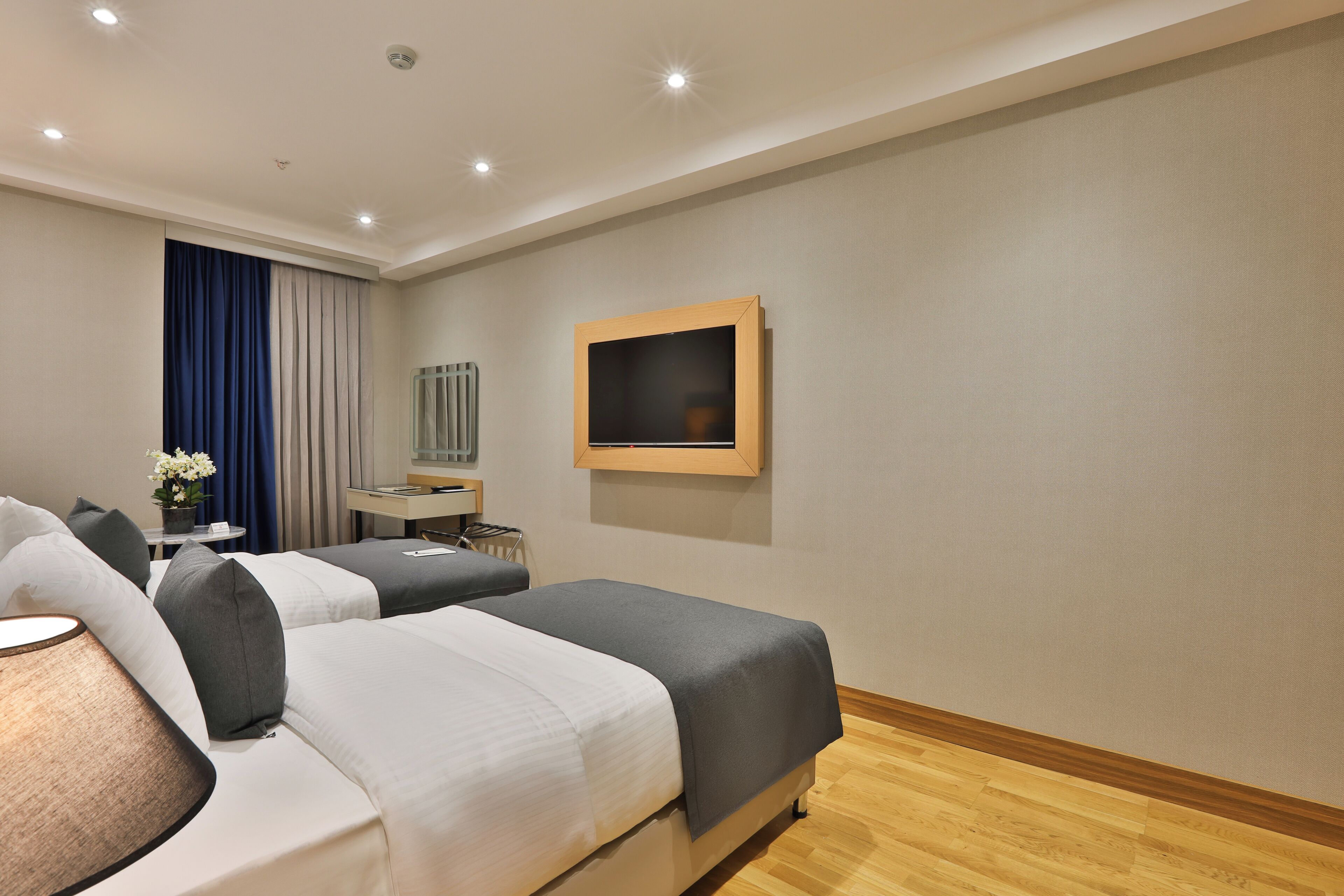 Quarto de hotel com duas camas, uma escrivaninha, uma cadeira, um espelho e uma televisão fixada na parede.