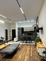 Uma sala de estar moderna com um sofá cinzento, uma mesa de centro de madeira e um banco cinzento. Há uma área de cozinha com uma mesa de jantar e cadeiras, um micro-ondas e uma chaleira. O quarto tem piso de madeira e uma janela grande com cortinas.