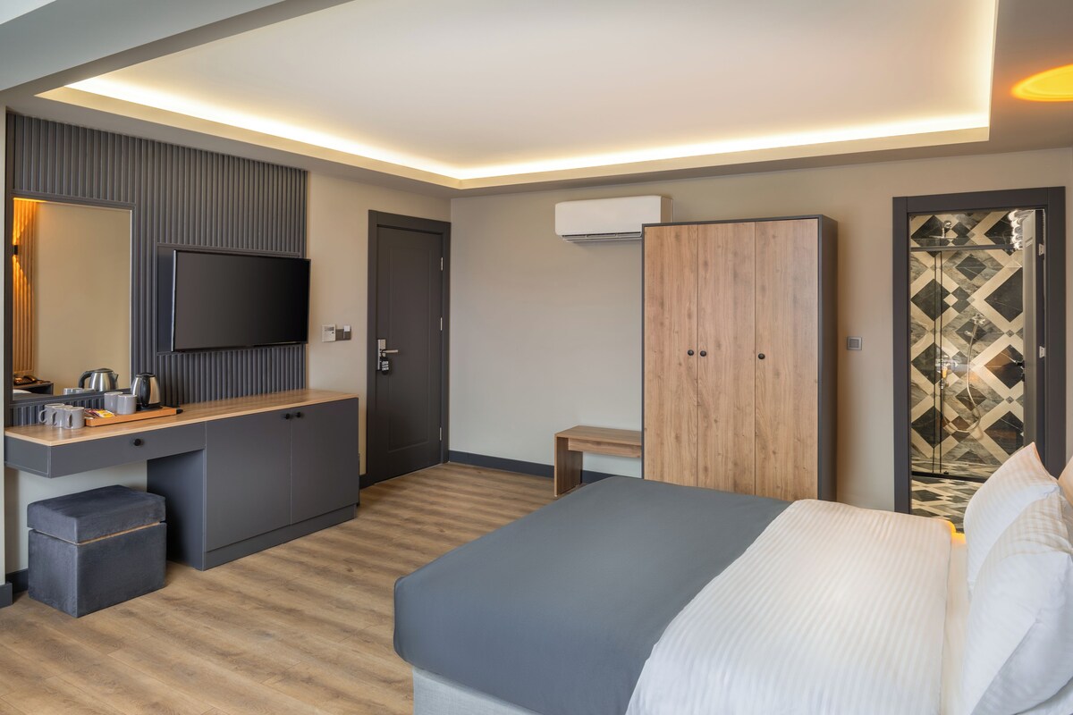 Quarto de hotel moderno com uma cama grande, uma televisão de tela plana na parede, um guarda-roupa de madeira e uma pequena escrivaninha com cadeira.