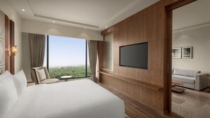 Quarto de hotel moderno com uma cama grande, televisão na parede, área de estar com uma cadeira e mesa, e vista da cidade através de janelas amplas.