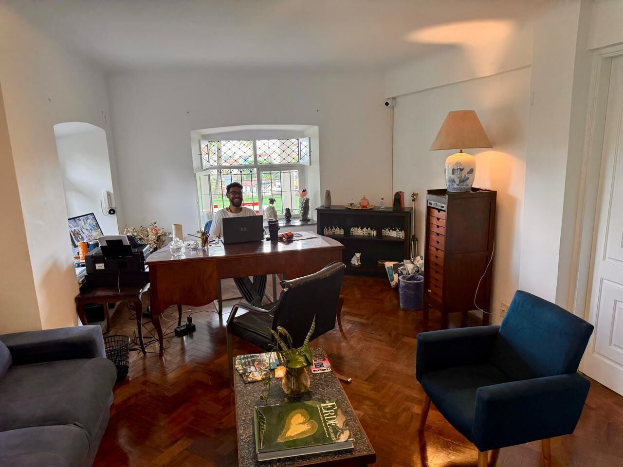 Uma sala de estar com piso de madeira, uma escrivaninha com um laptop, uma cadeira, um sofá e um abajur.