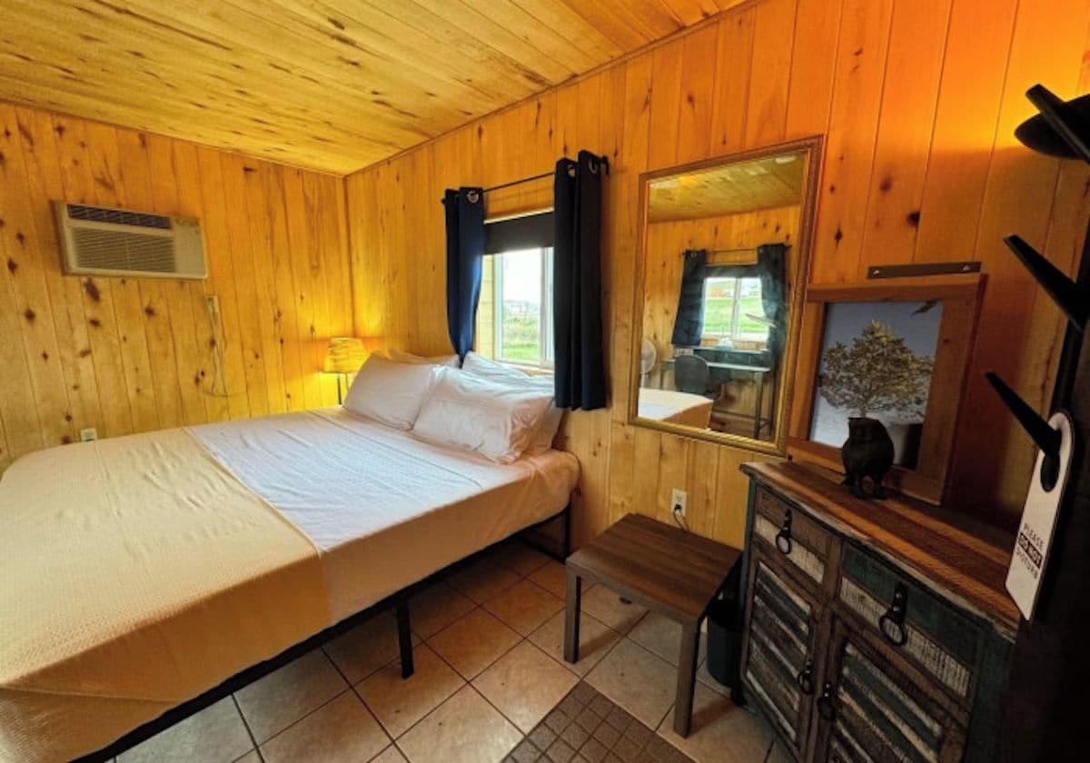 Quarto de madeira com uma cama, cômoda, espelho e janela com cortinas.