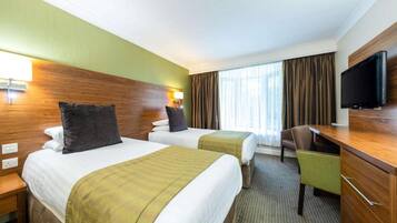 Quarto de hotel com duas camas, uma mesa, uma cadeira, uma televisão e uma janela com cortinas.