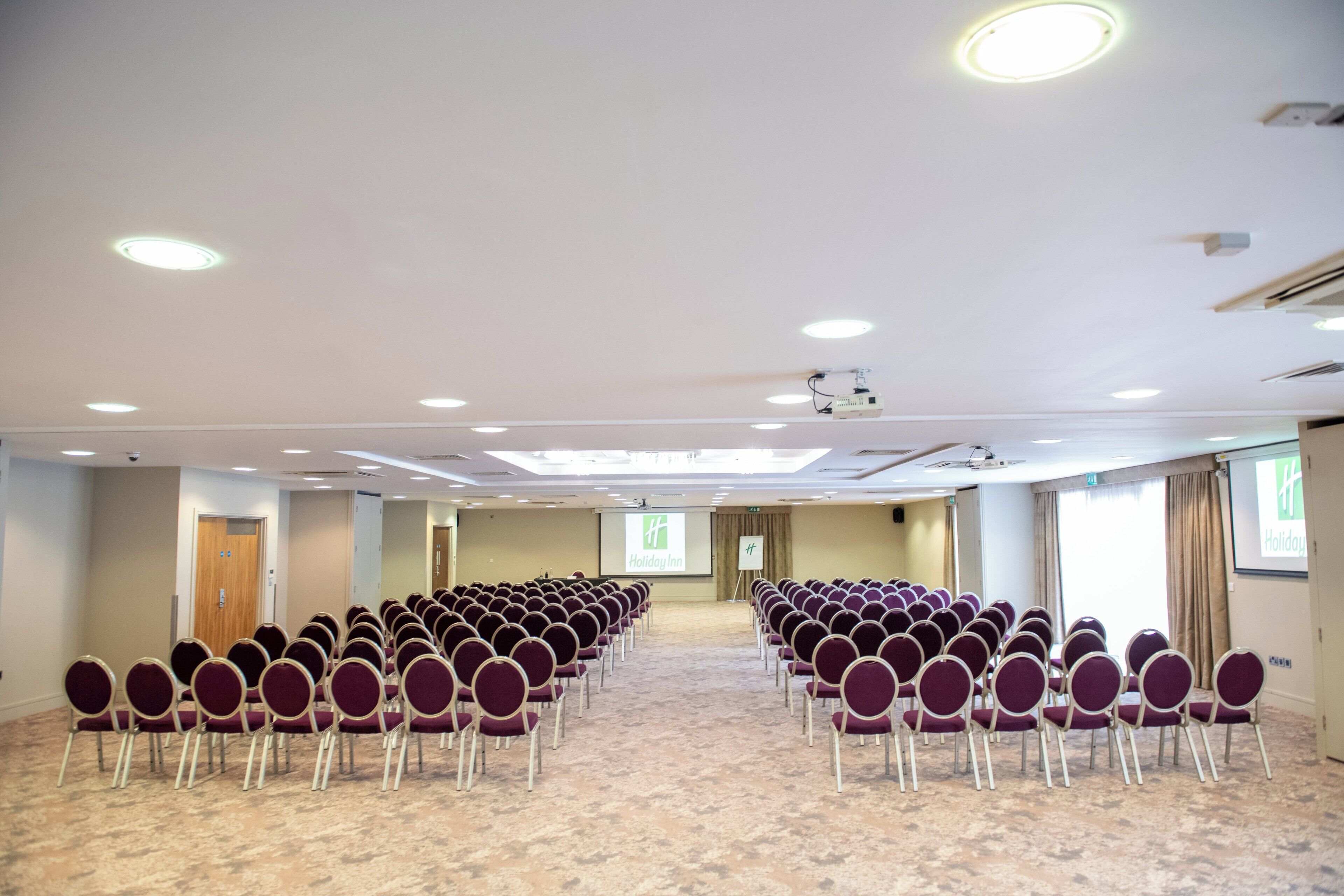 Sala de conferências com fileiras de cadeiras, projetor e tela exibindo 'Holiday Inn'.