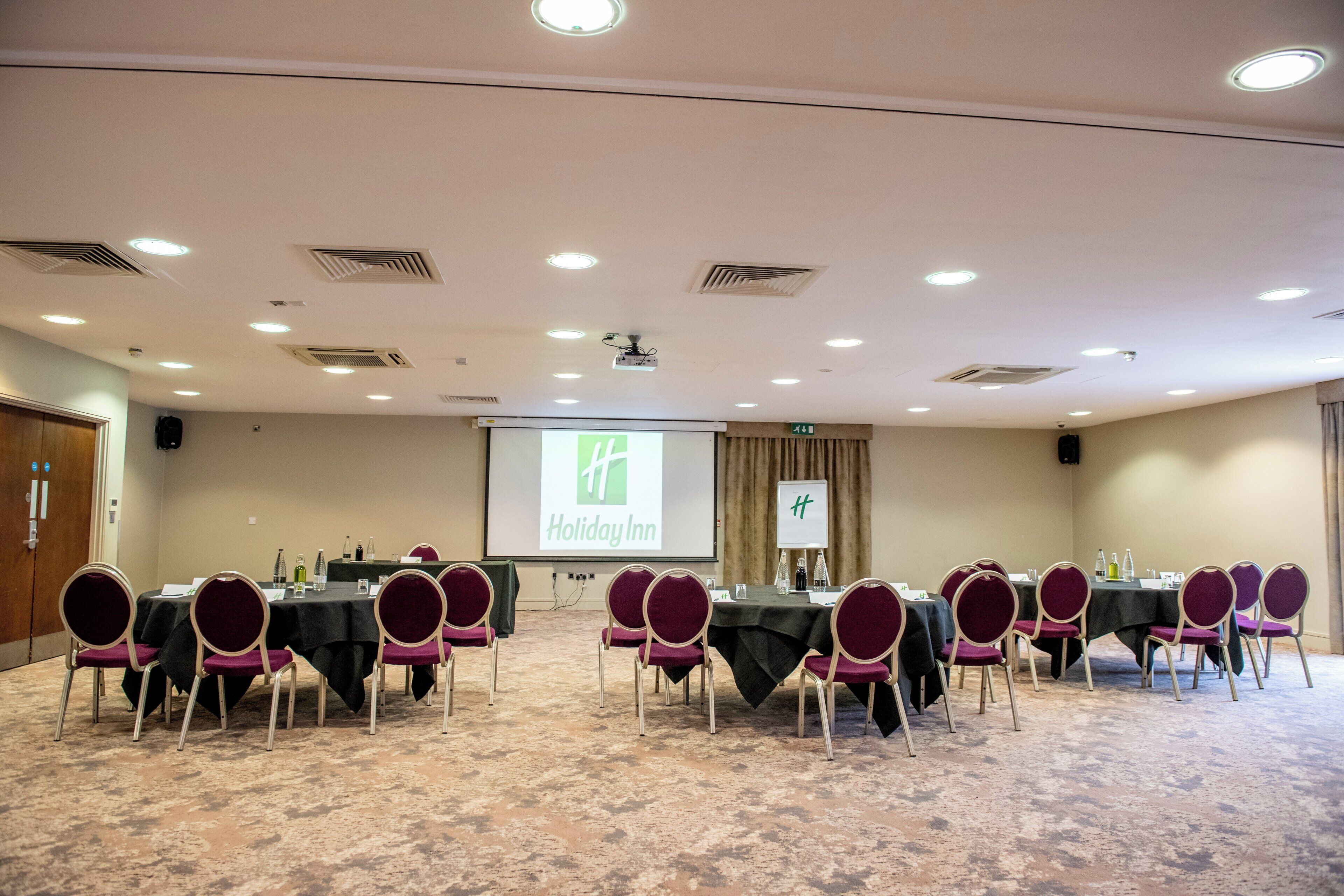 Uma sala de conferência com várias mesas e cadeiras, uma tela de projetor exibindo 'Holiday Inn' e piso coberto de carpete.