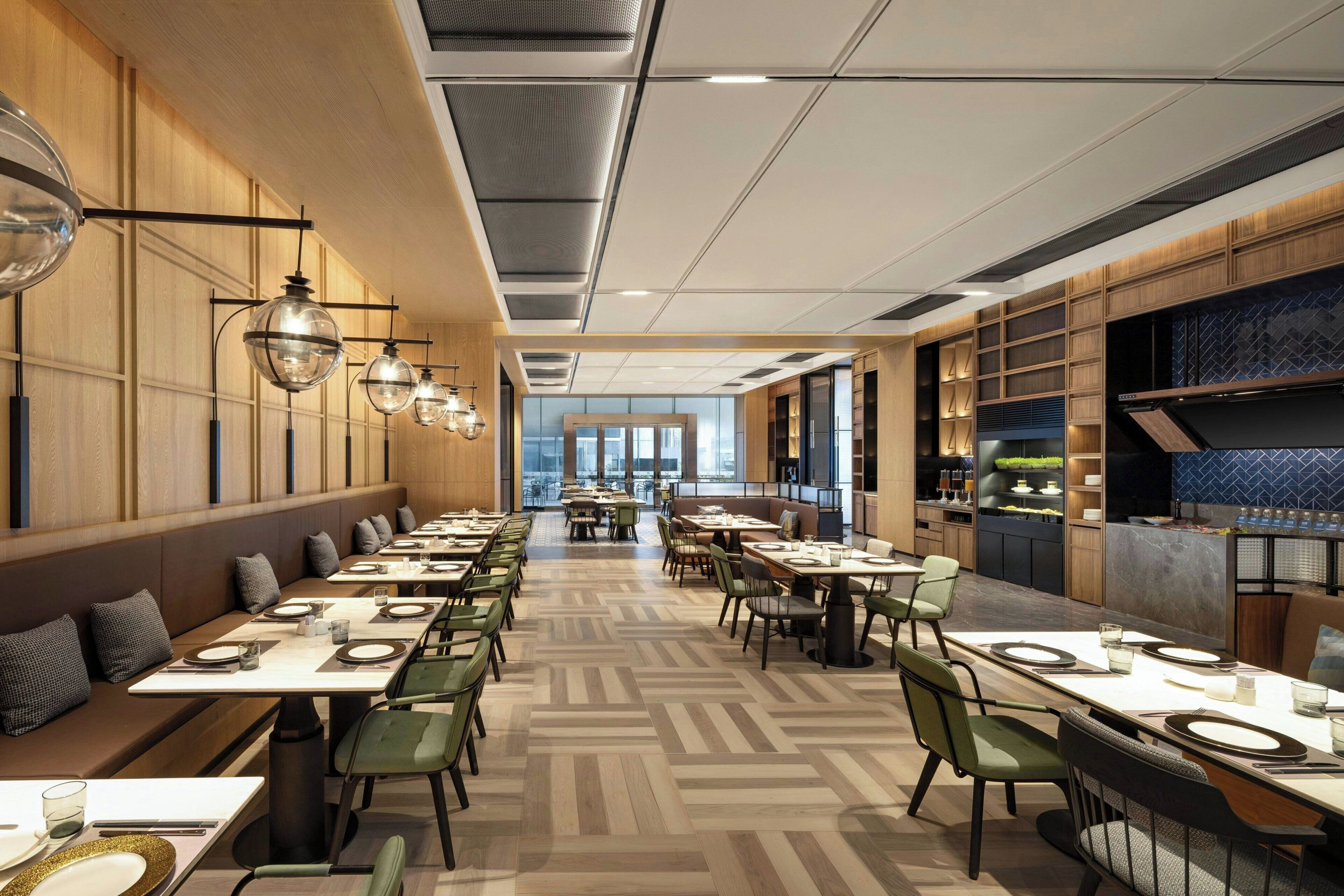 Interior de restaurante moderno com mesas grandes comunitárias, cadeiras verdes e luminárias pendentes.