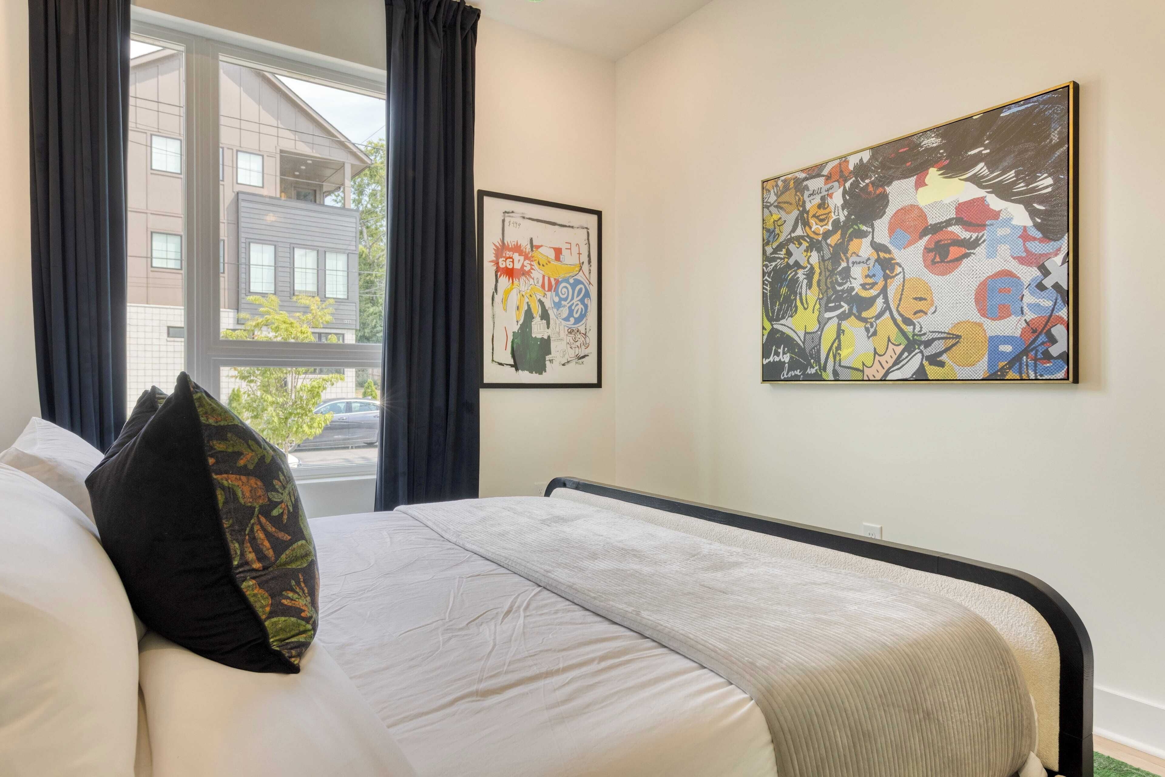 Quarto de hotel com uma cama, uma janela com cortinas e duas obras de arte emolduradas na parede.
