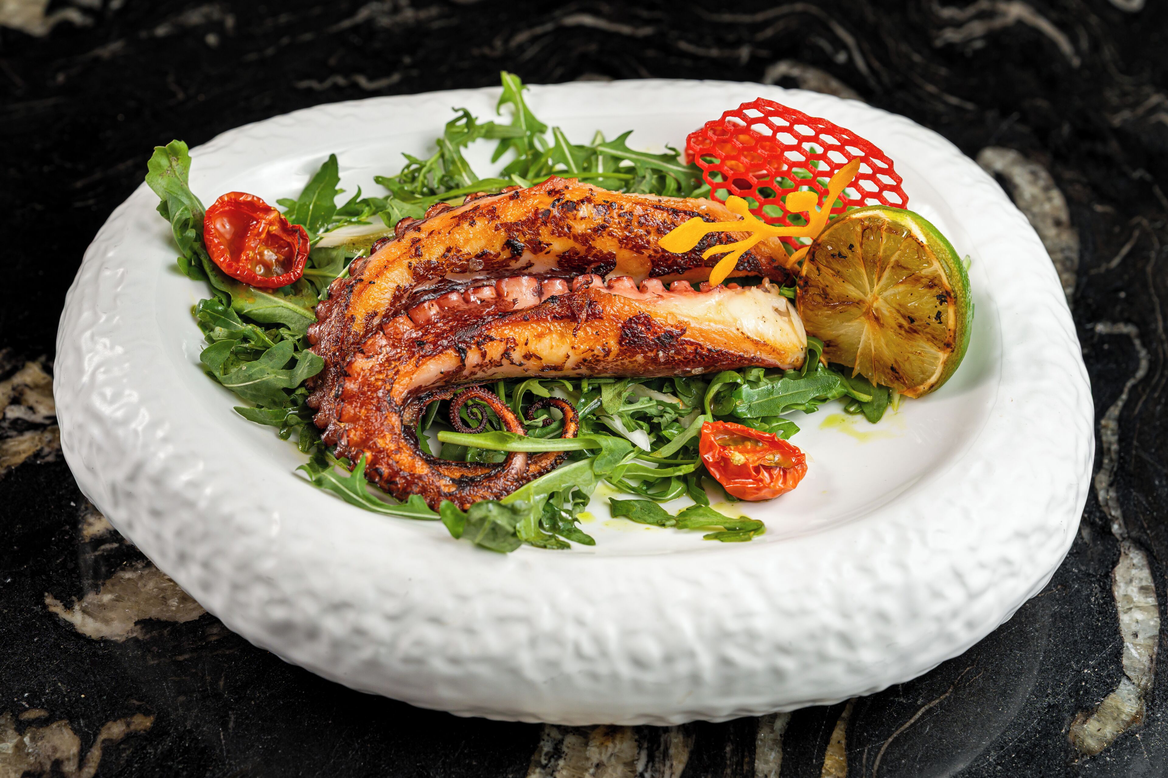 Um prato branco com polvo grelhado, tomates cereja e limão sobre uma superfície de mármore escuro.
