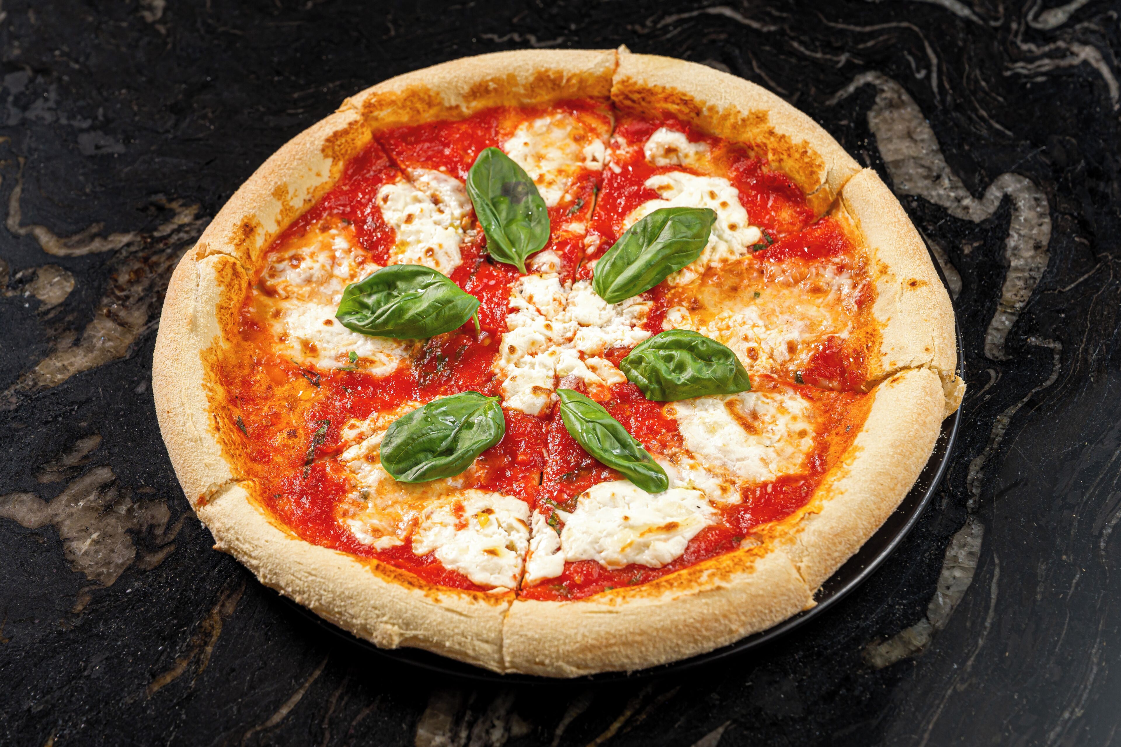 Pizza com molho de tomate, queijo e folhas de manjericão sobre uma superfície preta.