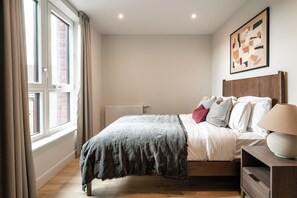Quarto com cama, mesa de cabeceira, abajur e janela com cortinas.