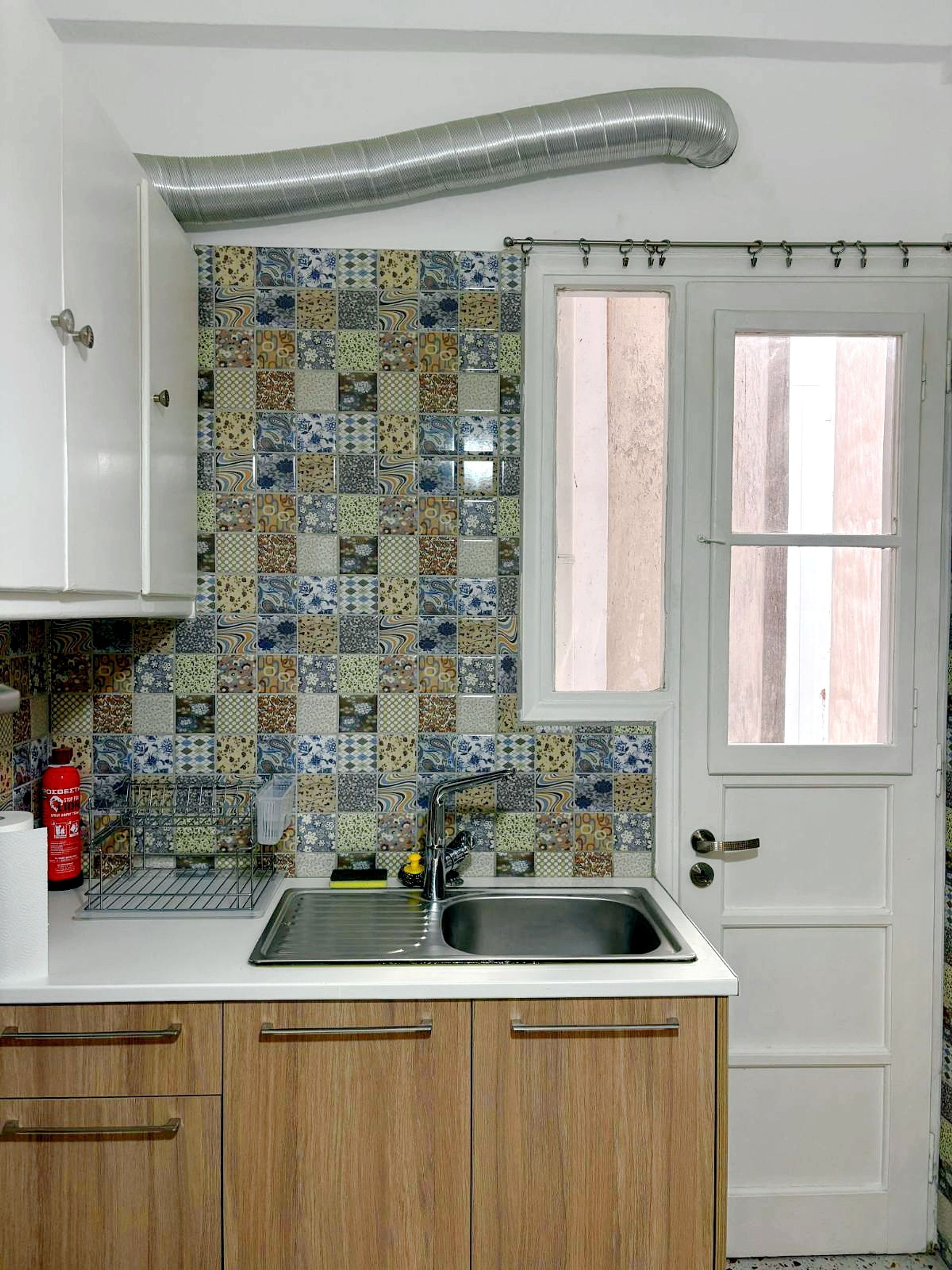 Uma cozinha com painel de azulejos, pia de aço inoxidável, armários de madeira e uma porta branca com janela.