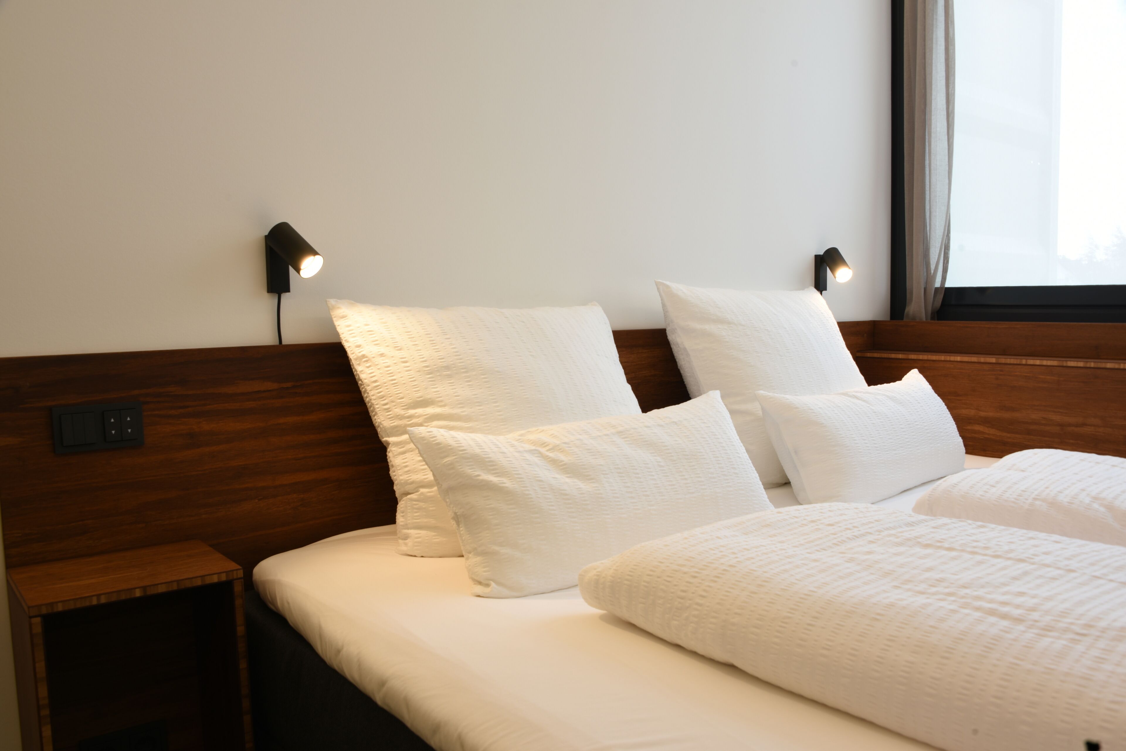 Cama bem arrumada com lençóis e almofadas brancas, cabeceira de madeira e duas luminárias de parede para leitura.