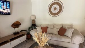 Sala de estar com um sofá, uma mesa de centro com um objeto decorativo de fibra natural, uma TV fixada na parede e um painel decorativo na parede.