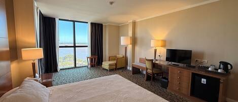 Quarto de hotel com uma cama grande, escrivaninha com televisão, uma cadeira e vista para o exterior.