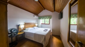Quarto com teto de madeira, cama com roupa de cama branca, mesa de cabeceira com um ventilador, janela com cortinas e TV fixada na parede.
