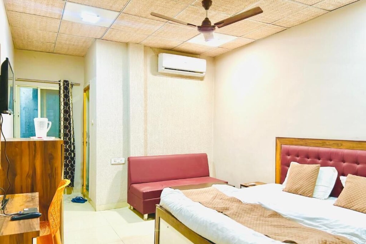 Quarto de hotel com cama, escrivaninha, cadeira, sofá vermelho, ventoinha no teto e ar condicionado.