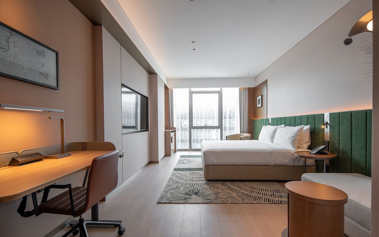 Quarto de hotel moderno com uma cama grande, uma escrivaninha com cadeira, uma mesinha de apoio e cabeceira verde.