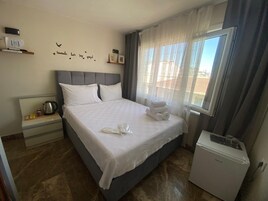 Quarto de hotel com cama, mesa de cabeceira com chaleira, mini-geladeira e janela com cortinas.
