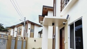 Um edifício residencial moderno de dois andares com fachada branca, varanda no andar superior e um pequeno jardim com plantas em vasos.