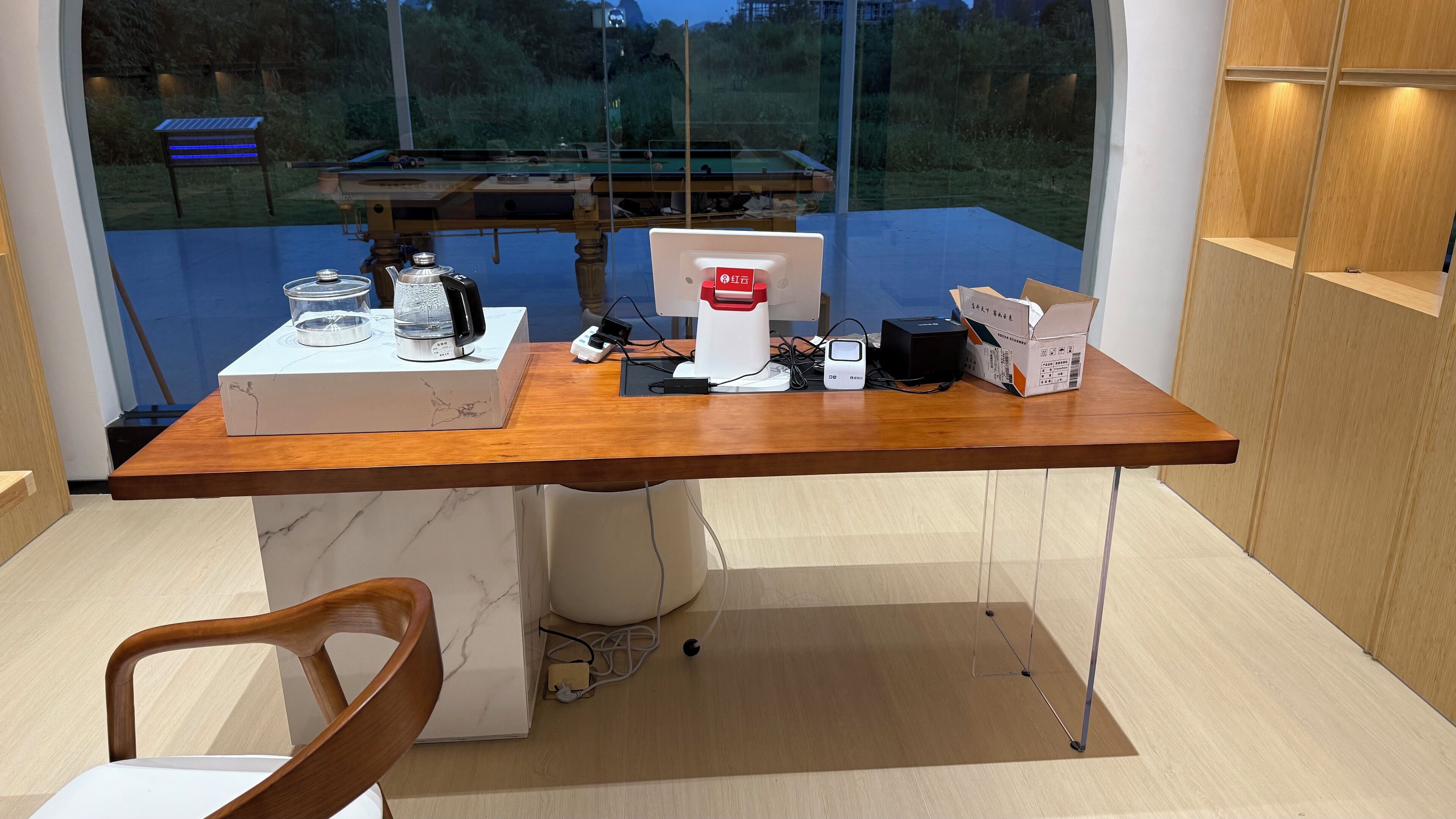 Uma secretária de madeira com um computador, uma chaleira e uma caixa, num quarto com vista para uma mesa de bilhar e vegetação lá fora.