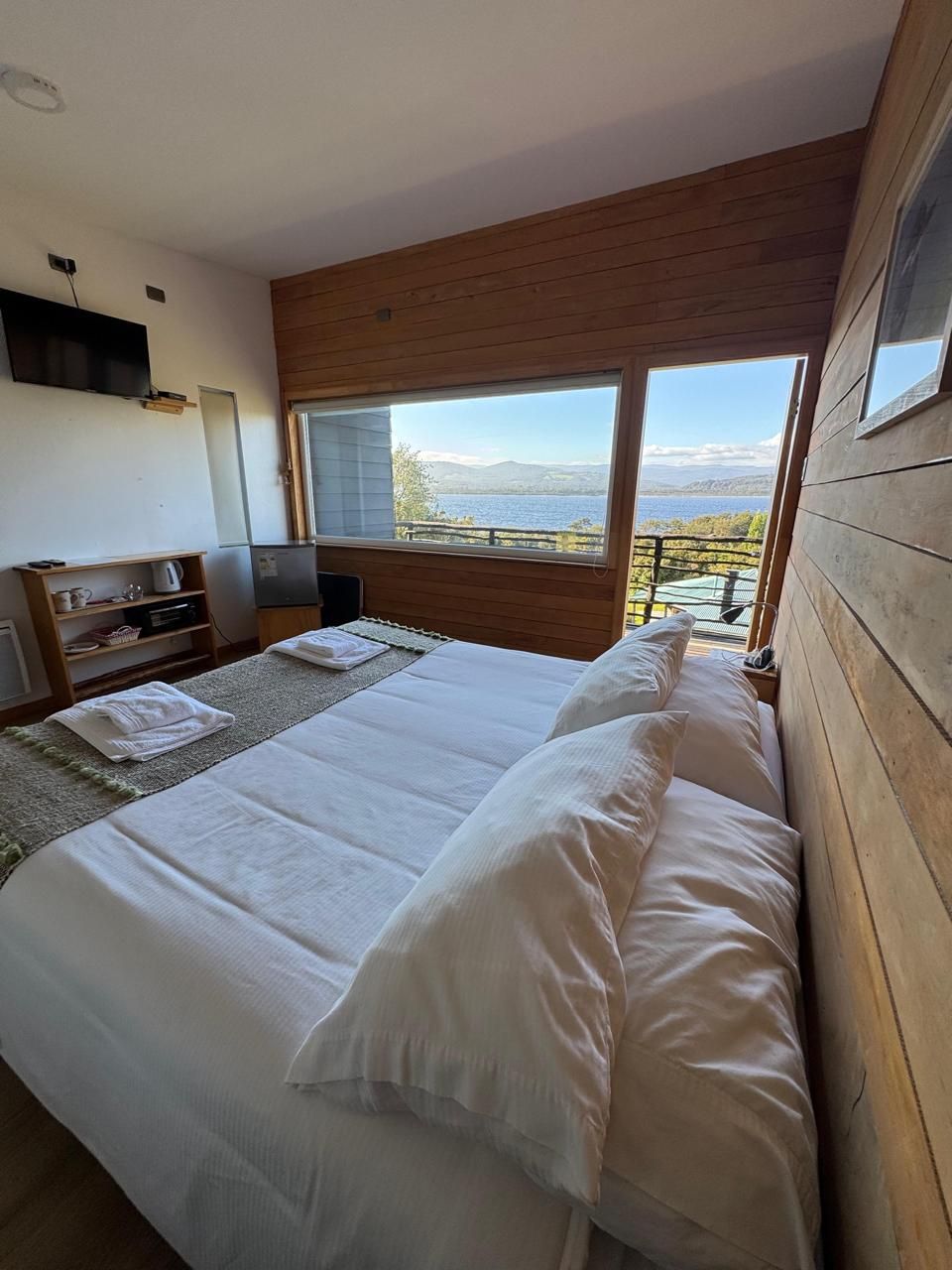 Quarto com uma cama grande, paredes de madeira, televisão e vista para um lago.