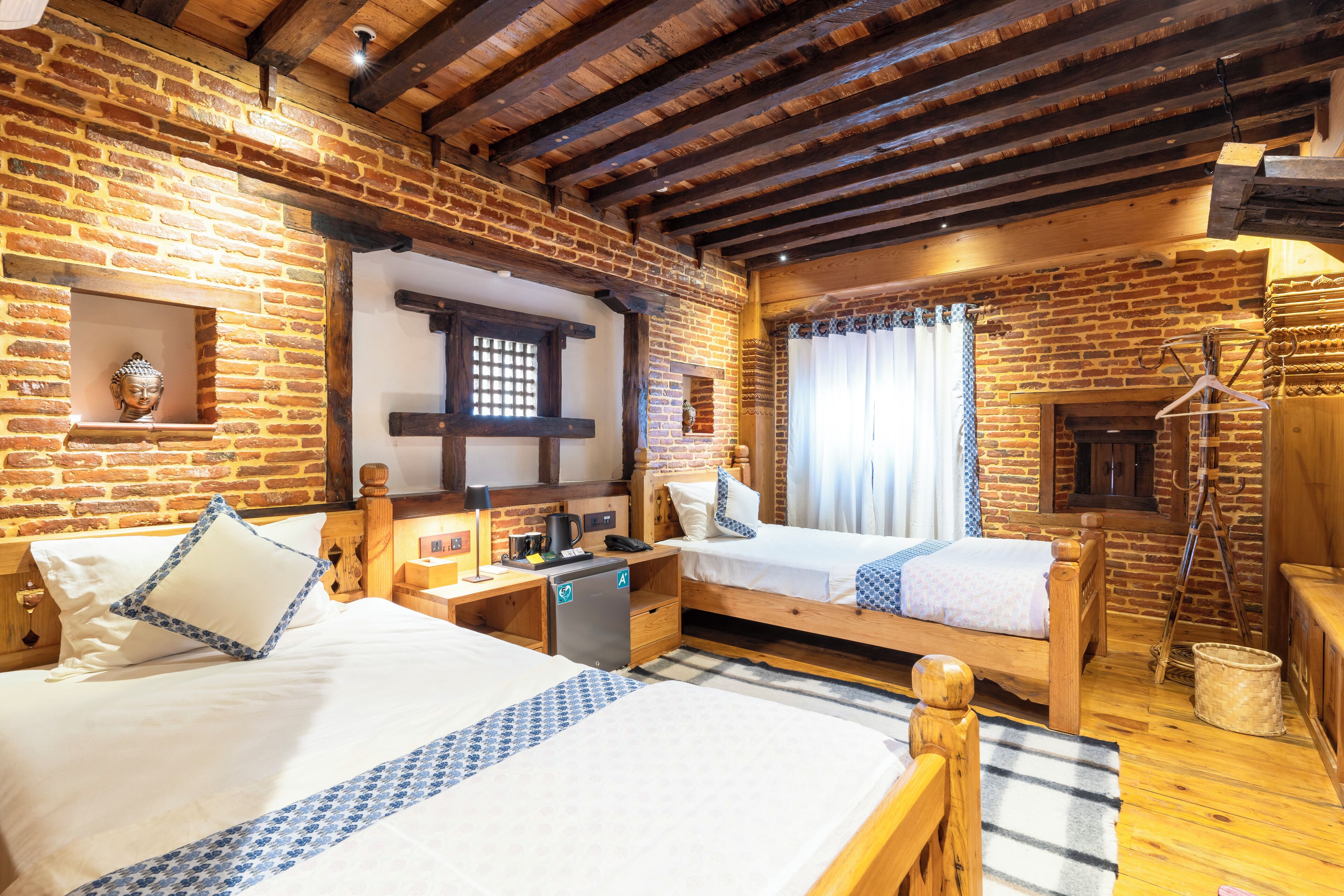 Quarto rústico com paredes de tijolo aparente, vigas de madeira, cama com cabeceira de madeira, pequena escrivaninha com abajur e um cabideiro.
