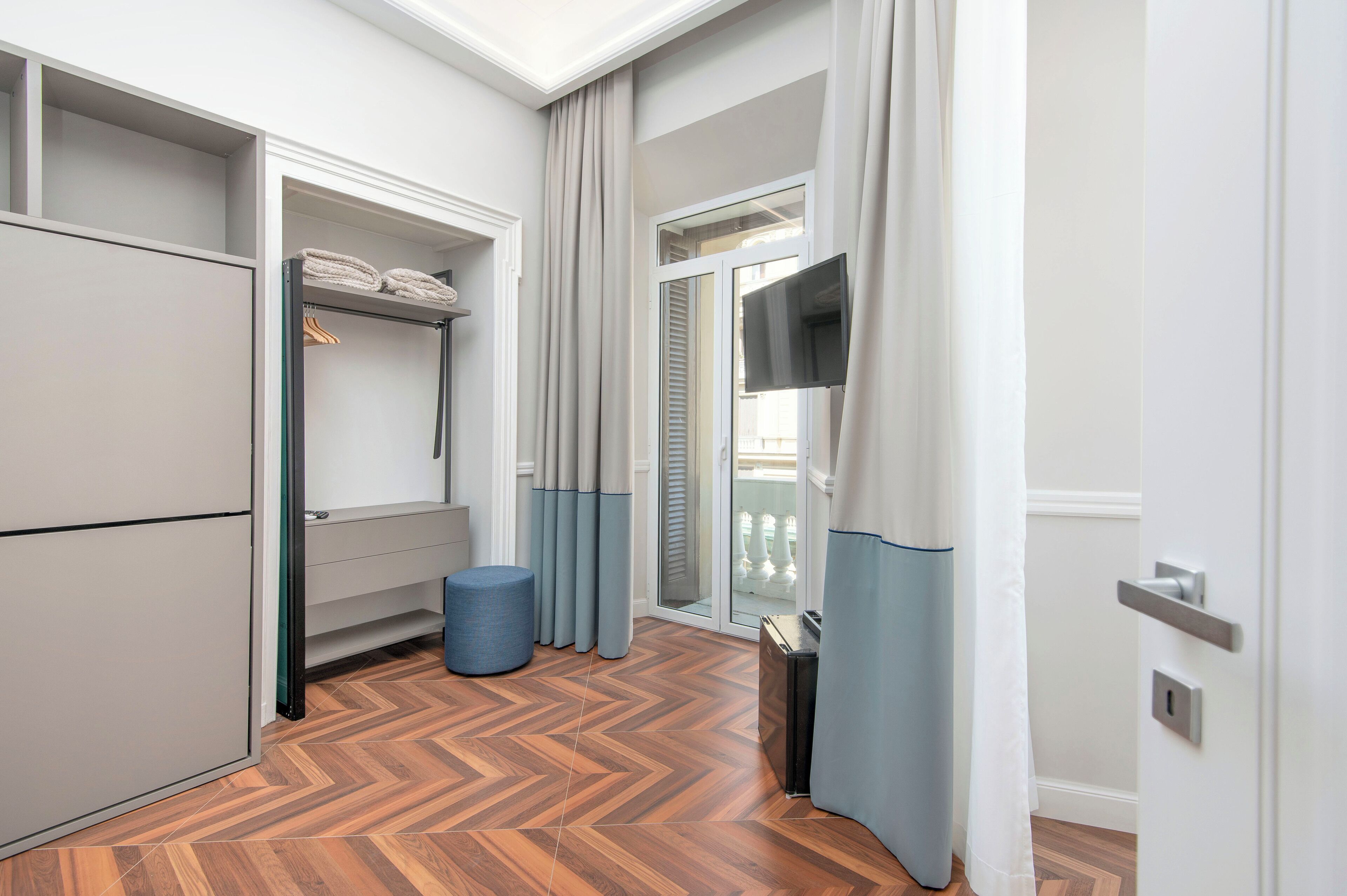 Quarto de hotel moderno com piso de madeira em padrão herringbone, armário embutido, área de estar com um banquinho azul e porta de vidro deslizante para uma varanda.