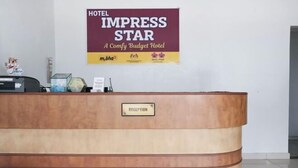 Uma mesa de recepção com uma placa que diz "HOTEL IMPRESS STAR A Comfy Budget Hotel" e a etiqueta "RECEPÇÃO".
