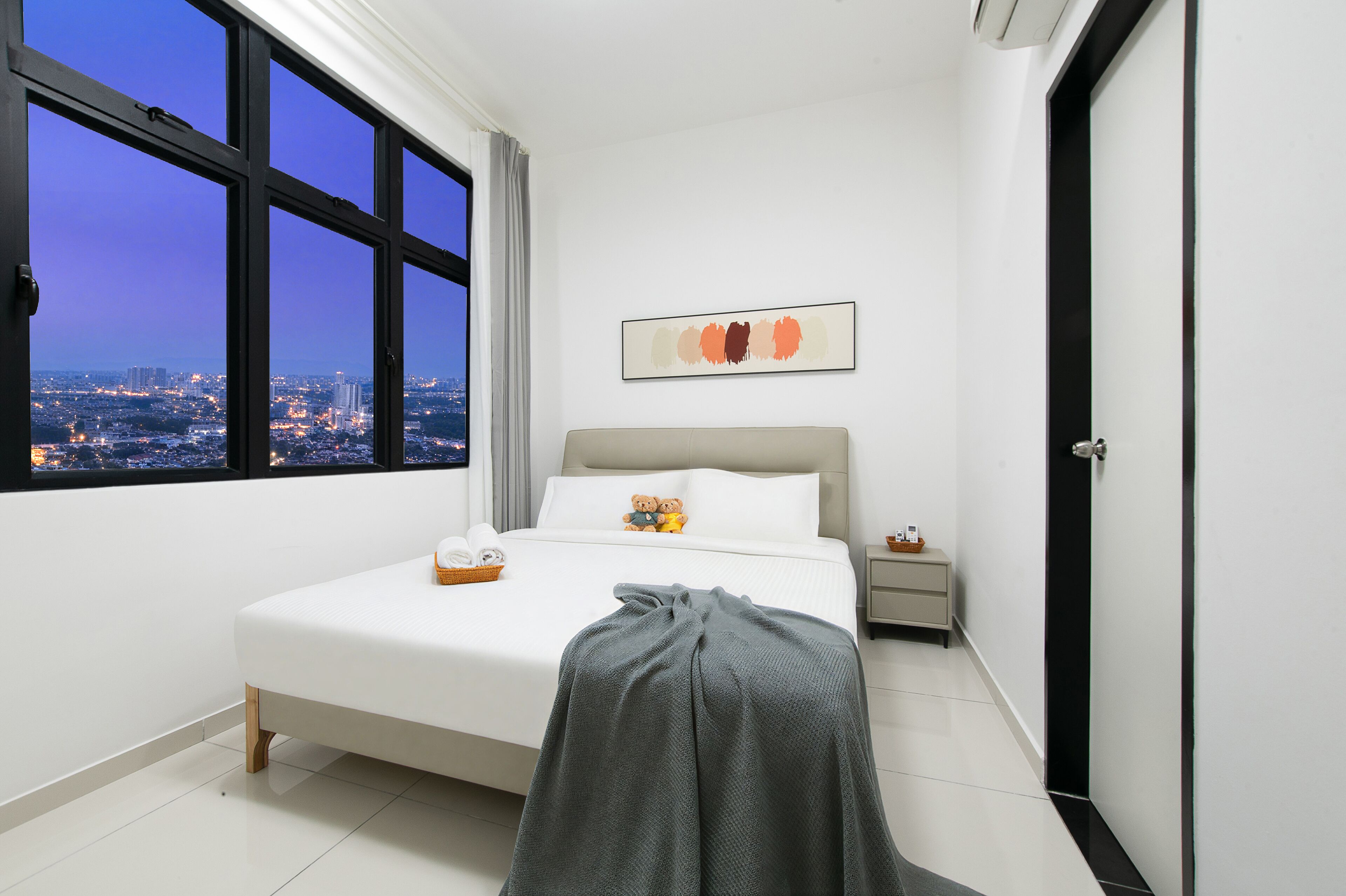 Quarto de hotel moderno com uma grande janela que oferece vista para a cidade, uma cama bem arrumada, uma mesinha de cabeceira com abajur e uma obra de arte abstrata na parede.