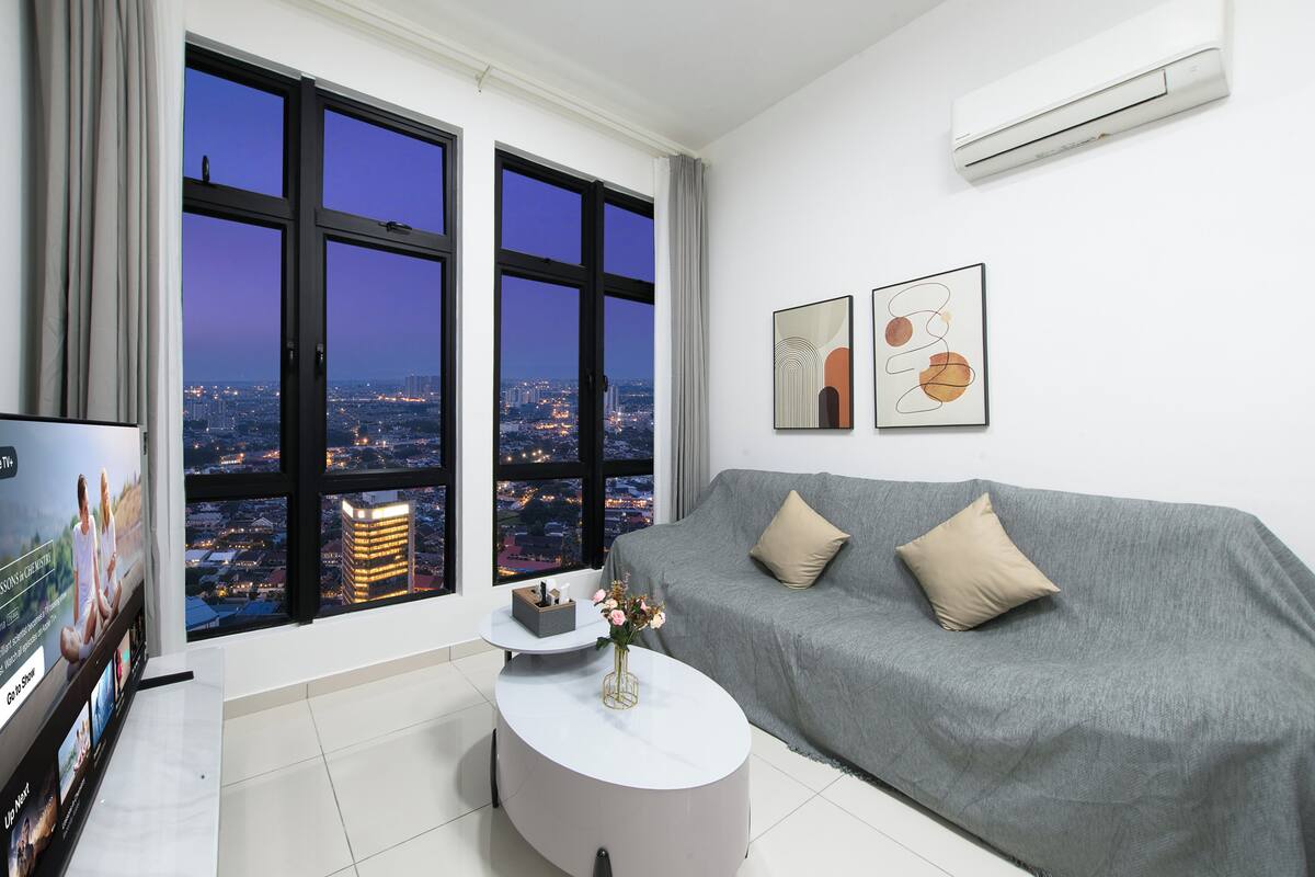 Uma sala de estar moderna com um sofá cinzento, uma mesa de centro redonda e uma TV à esquerda. Grandes janelas proporcionam uma vista da cidade ao anoitecer.