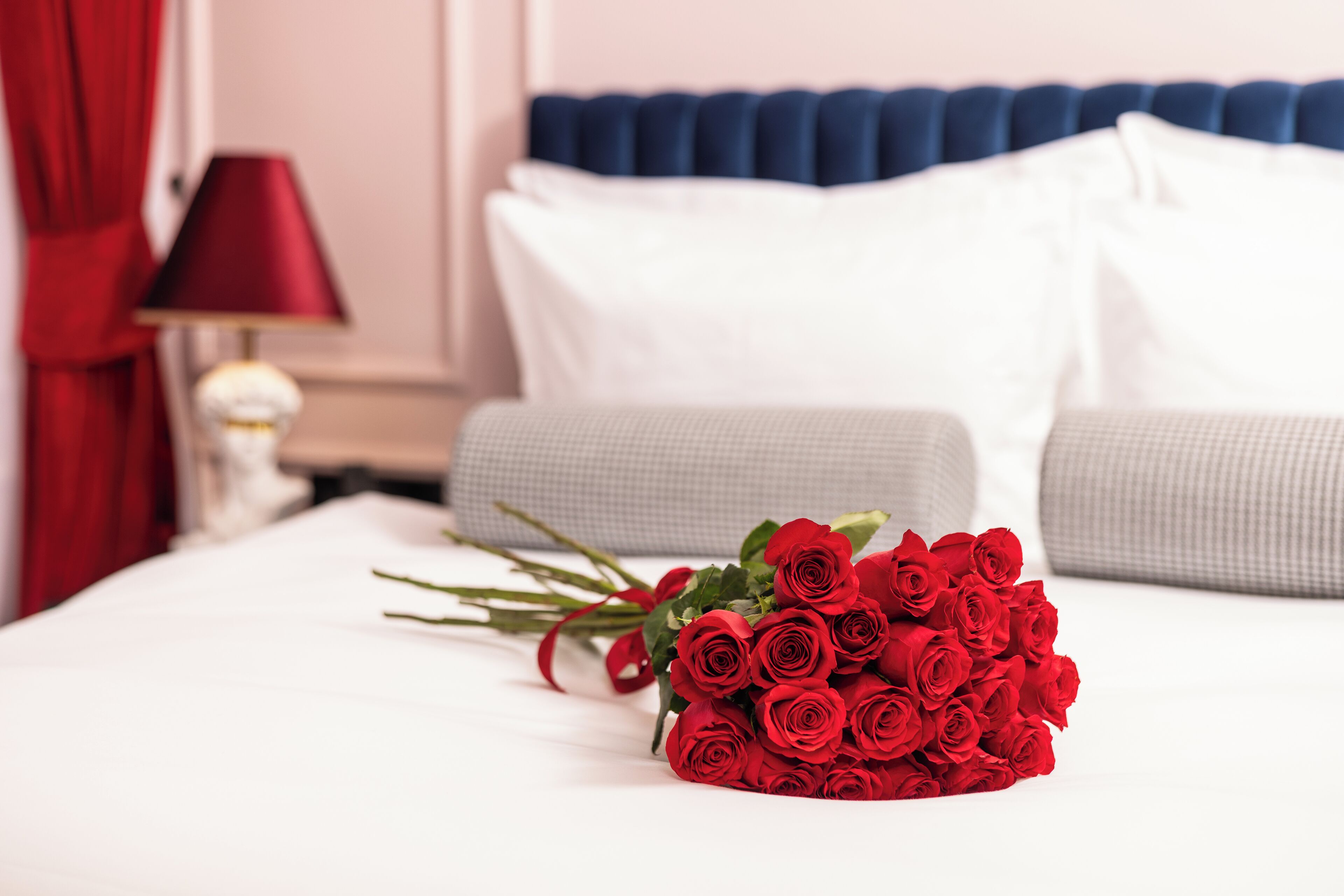 Uma cama com lençóis brancos, um buquê de rosas vermelhas e um abajur vermelho em uma mesa de cabeceira.