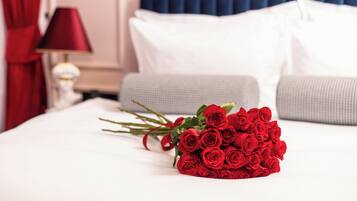 Uma cama com lençóis brancos, um buquê de rosas vermelhas e um abajur vermelho em uma mesa de cabeceira.