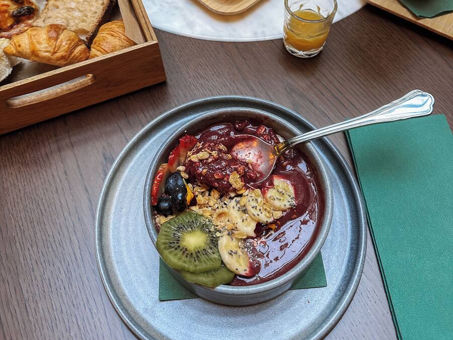Mesa de café da manhã com uma tigela de frutas e granola, uma bandeja de doces e pequenos recipientes de condimentos.