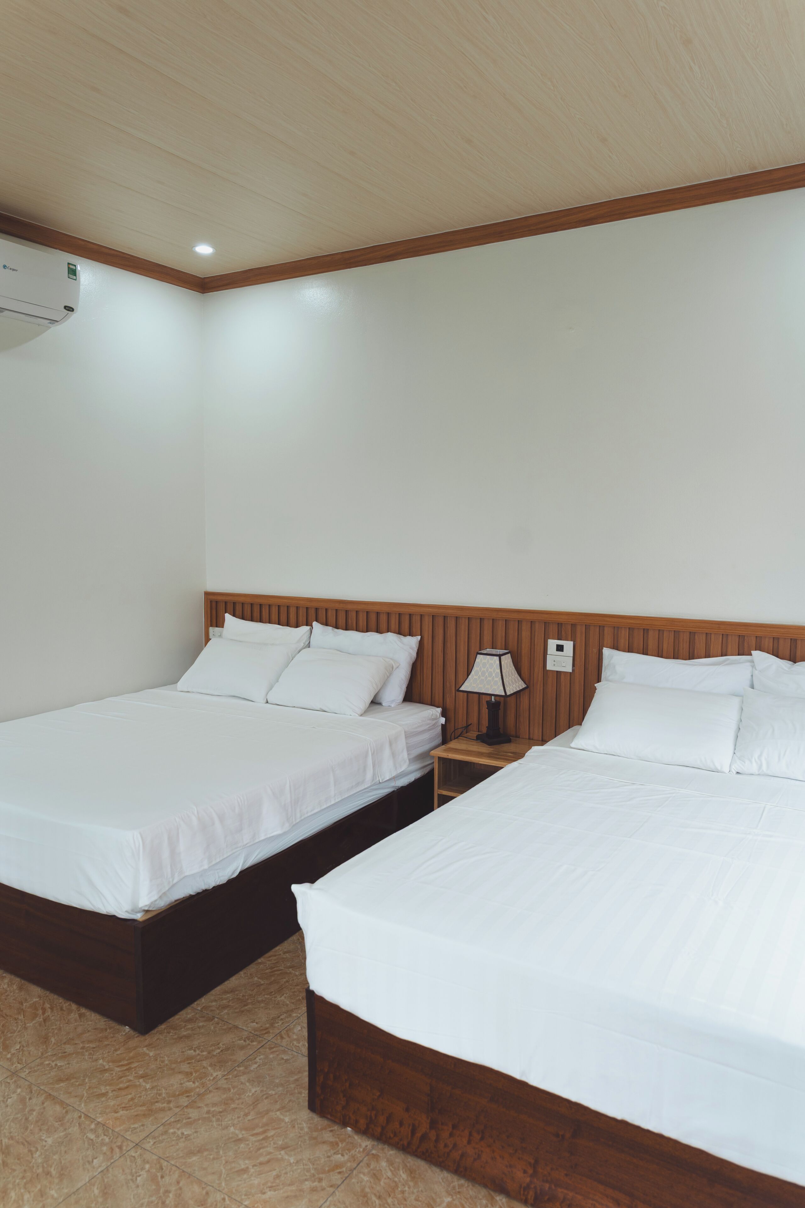 Quarto de hotel com duas camas, cabeceira de madeira, mesa de cabeceira com abajur e unidade de ar condicionado.