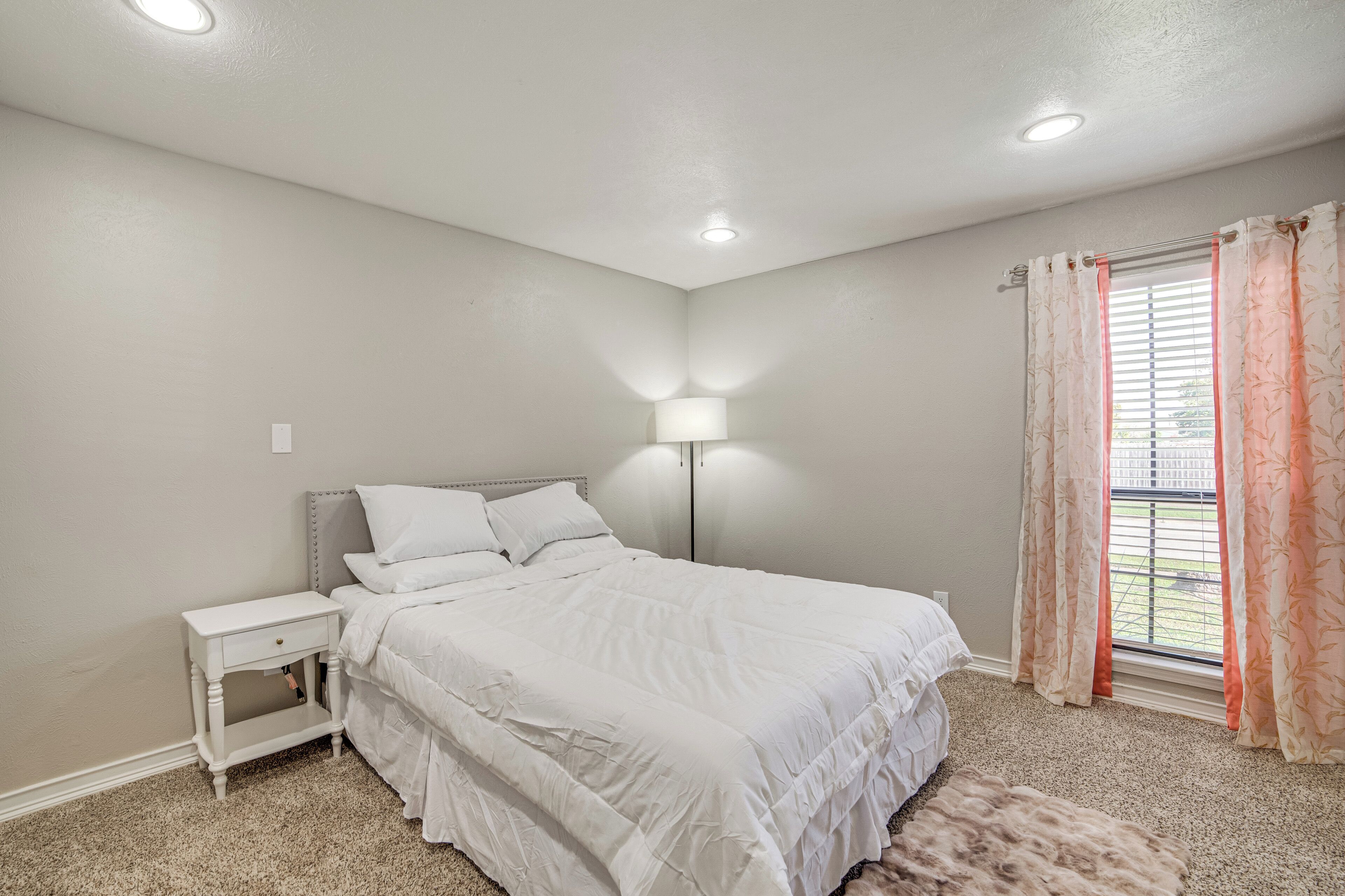 Quarto com cama, mesa de cabeceira, luminária e janela com cortinas.
