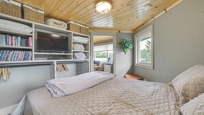 Quarto com cama, estantes com livros e cestos, televisão e janela com vista para árvores.