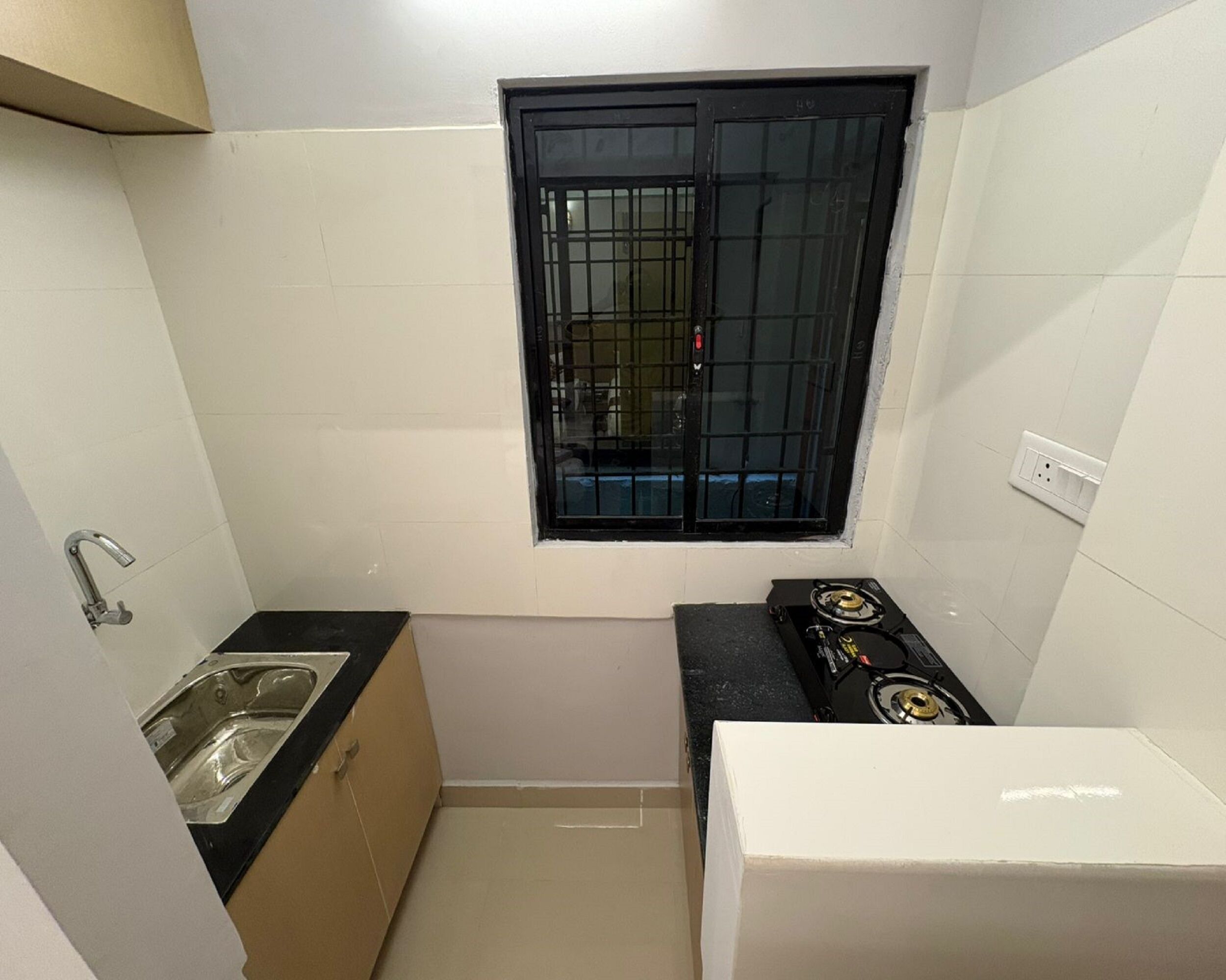 Uma cozinha pequena e compacta, com paredes revestidas de azulejos brancos, uma janela preta com grades de metal, fogão a gás e uma pia.
