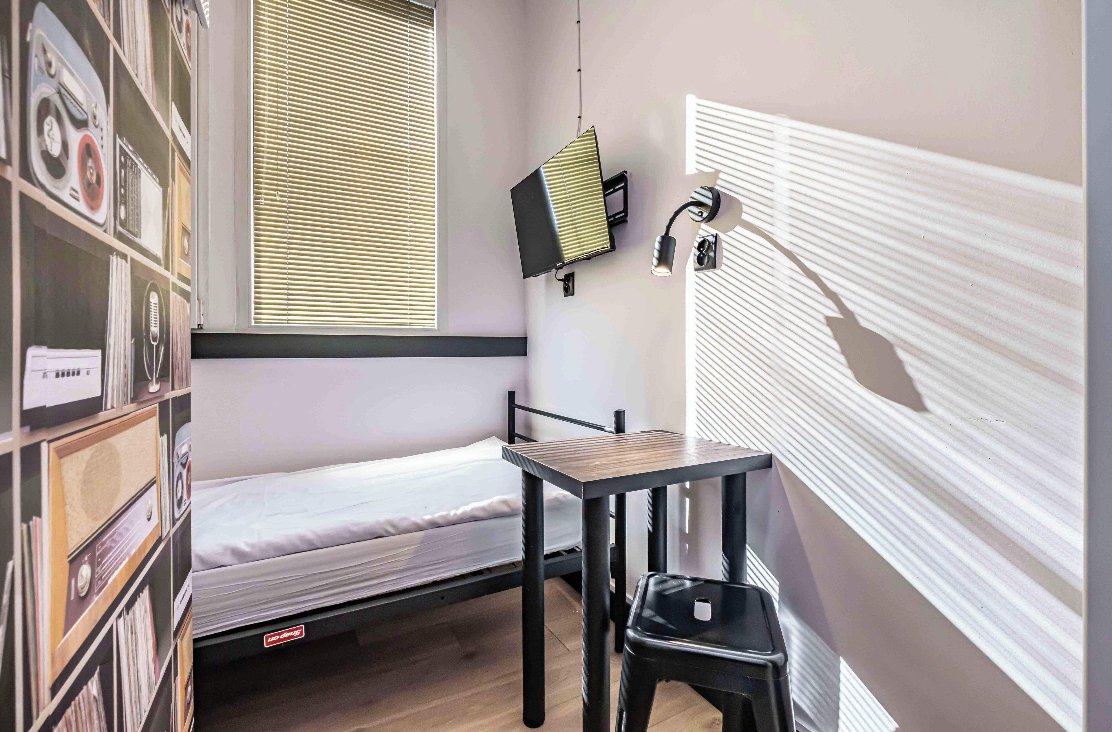 Quarto compacto com uma cama de solteiro, uma mesinha, uma cadeira, estante e televisão na parede.