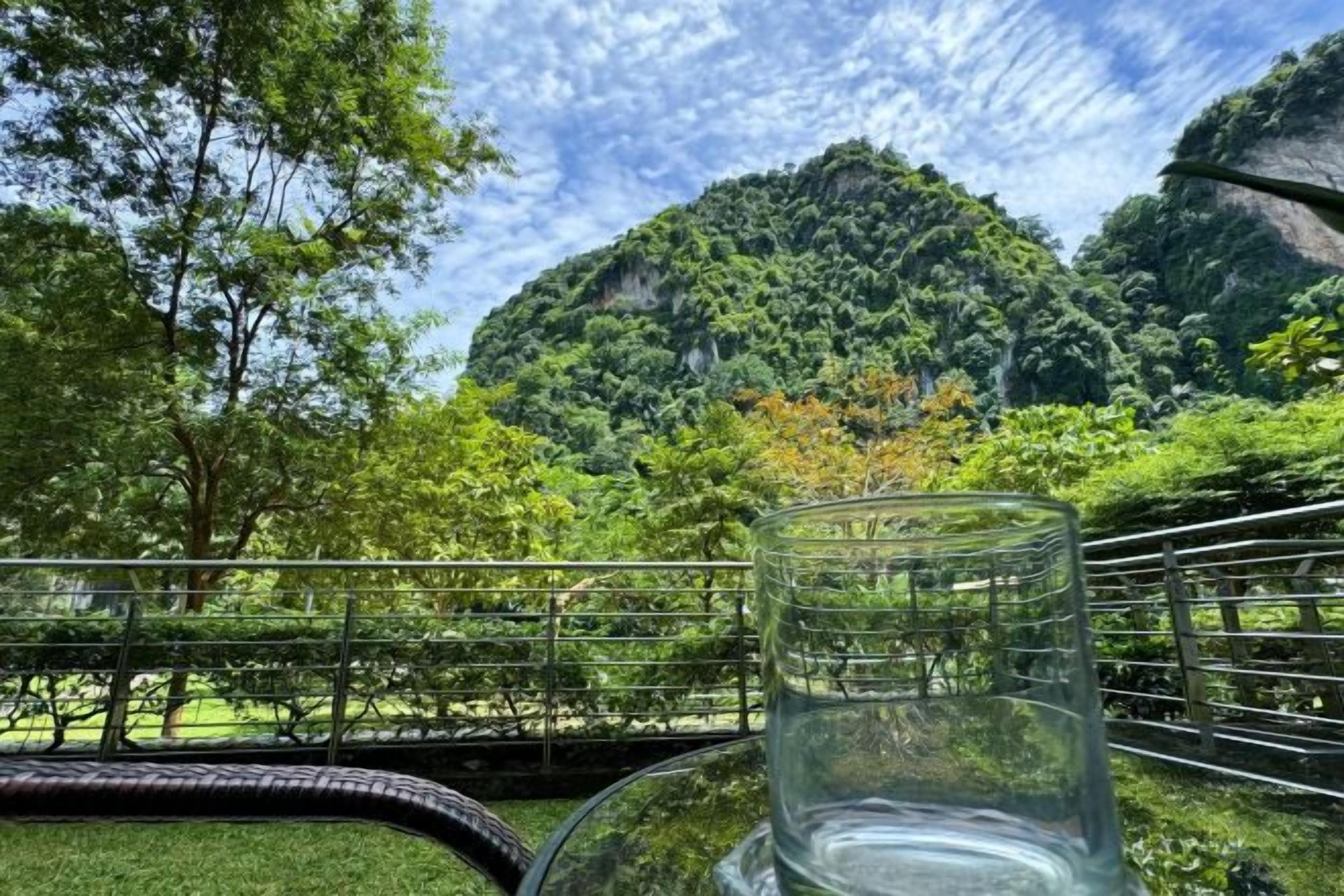 Um copo de água sobre uma mesa com corrimão metálico, com vista para uma paisagem verde exuberante e montanhas ao fundo.